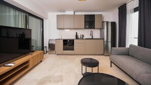 Uma sala moderna com televisão de tela plana, sofá, mesa redonda pequena e área de cozinha com armários e eletrodomésticos.