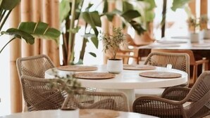 Uma área de refeições com mesas redondas, cadeiras de vime, luminárias pendentes e plantas verdes exuberantes.