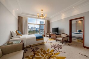Quarto de hotel com cama, televisão, área de estar com cadeiras, janela grande com vista para a cidade e um candelabro.