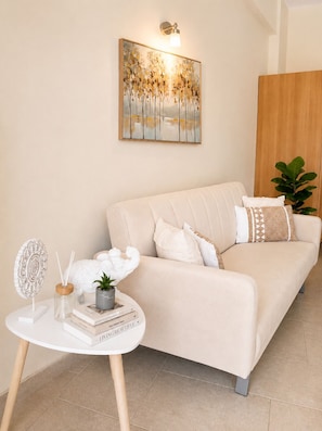 Uma sala de estar com um sofá bege, uma pequena mesa branca com objetos decorativos, uma luminária na parede e uma pintura.