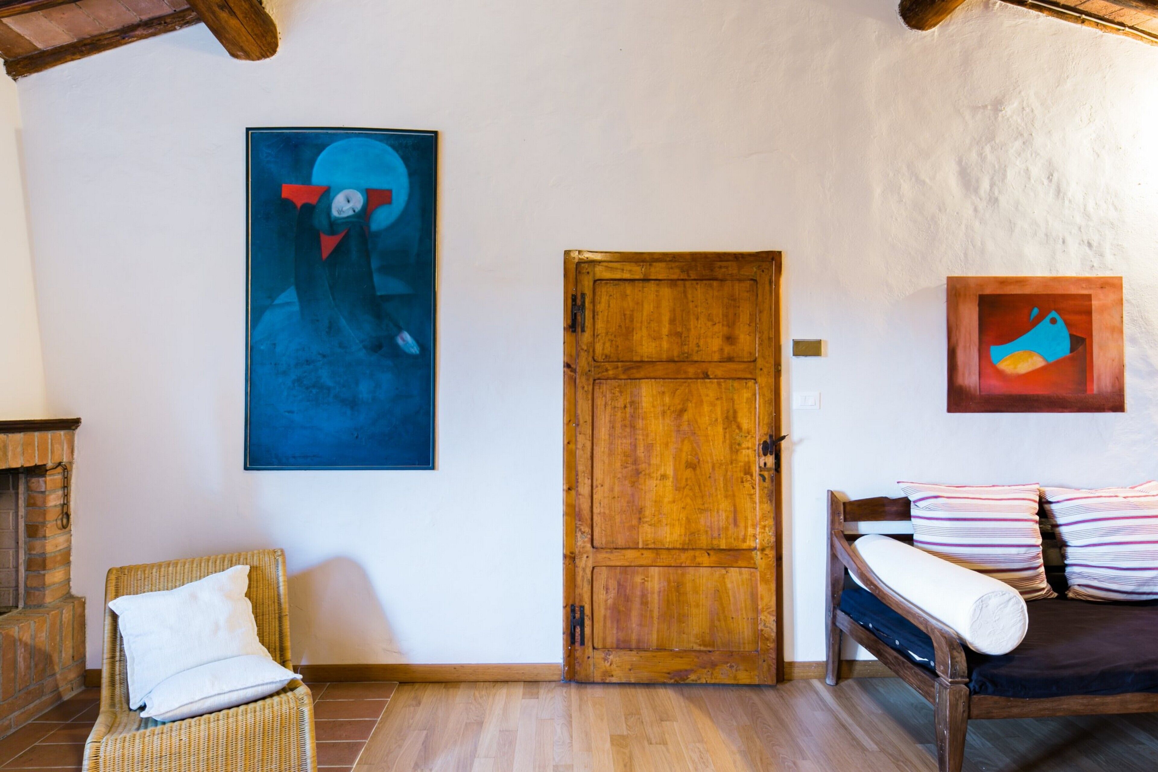 Um quarto com porta de madeira, uma cadeira de vime com almofada branca, uma cama com travesseiros listrados e duas pinturas abstratas na parede.