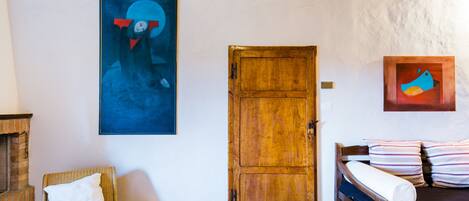 Um quarto com porta de madeira, uma cadeira de vime com almofada branca, uma cama com travesseiros listrados e duas pinturas abstratas na parede.