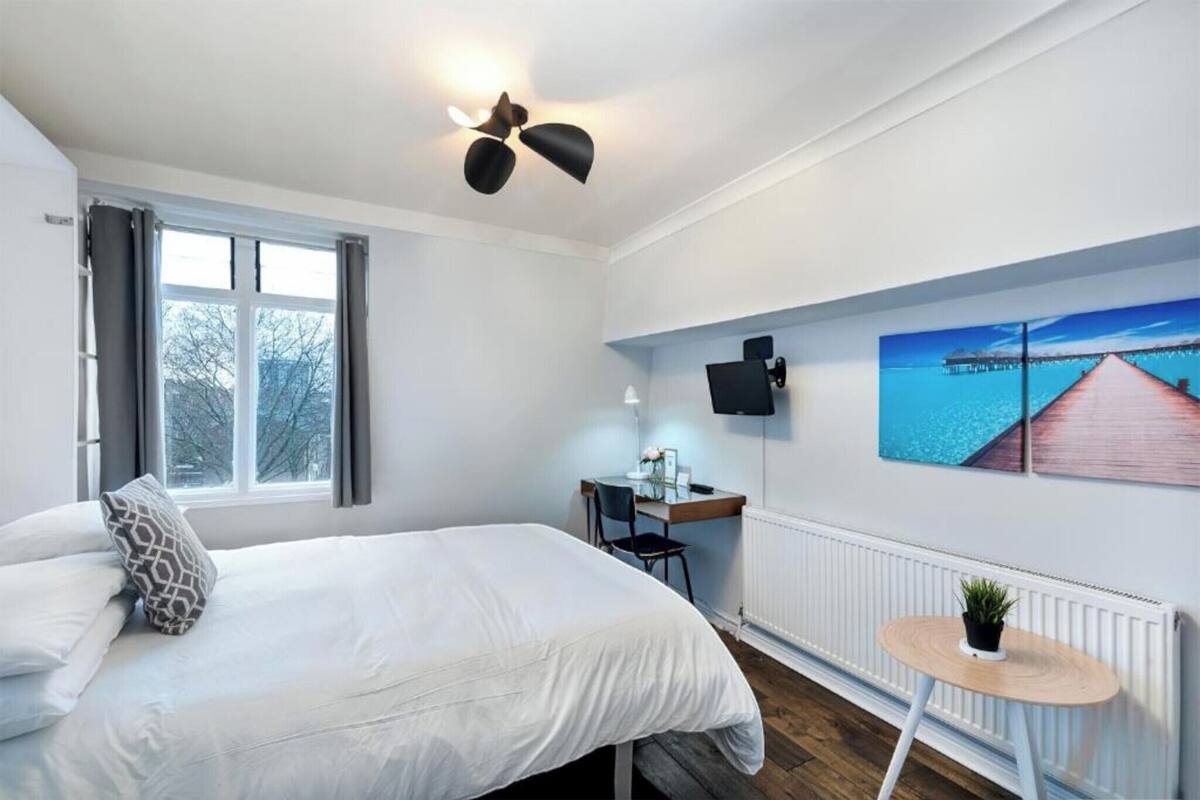 Quarto com cama, escrivaninha, cadeira, mesinha com uma planta, televisão e um quadro na parede.