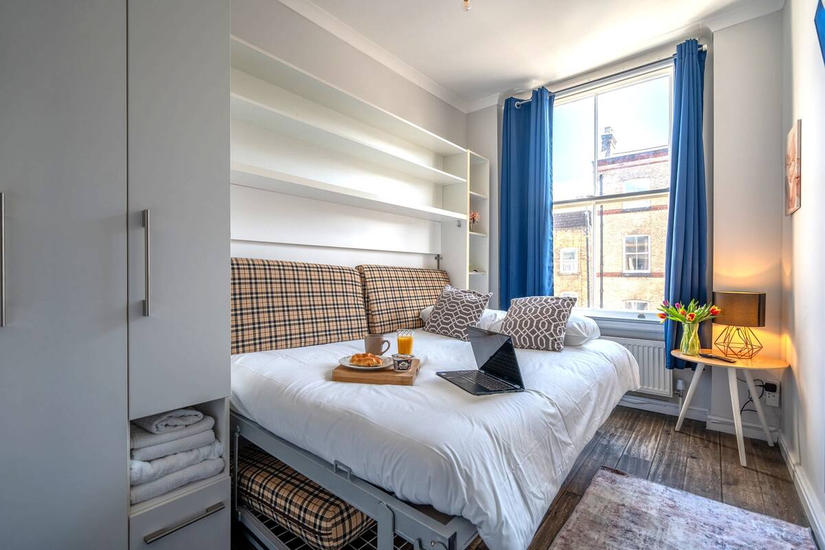 Quarto bem arrumado com cama, um laptop, uma bandeja de café da manhã e uma janela com cortinas.