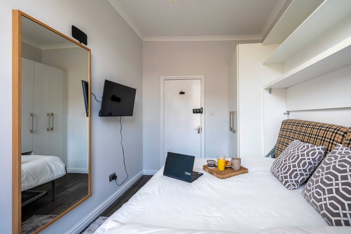 Quarto com uma cama, um laptop, uma TV, um espelho e uma porta.