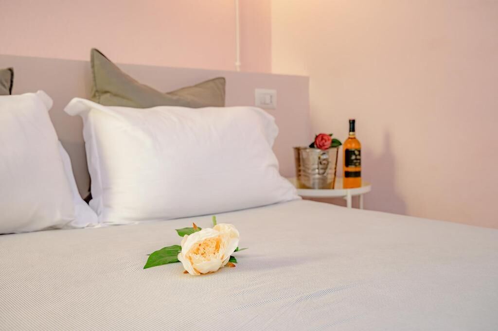Cama bem arrumada com roupa de cama branca, uma única rosa branca e uma garrafa de bebida sobre uma pequena mesa ao lado.