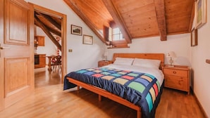 Quarto com teto de madeira, cama com colcha colorida, mesa de cabeceira e porta de madeira que dá para outro cômodo.