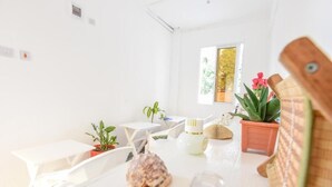 Um quarto claro com paredes brancas, uma janela com vista, uma mesa com uma concha, um vaso de planta e uma cadeira de madeira.