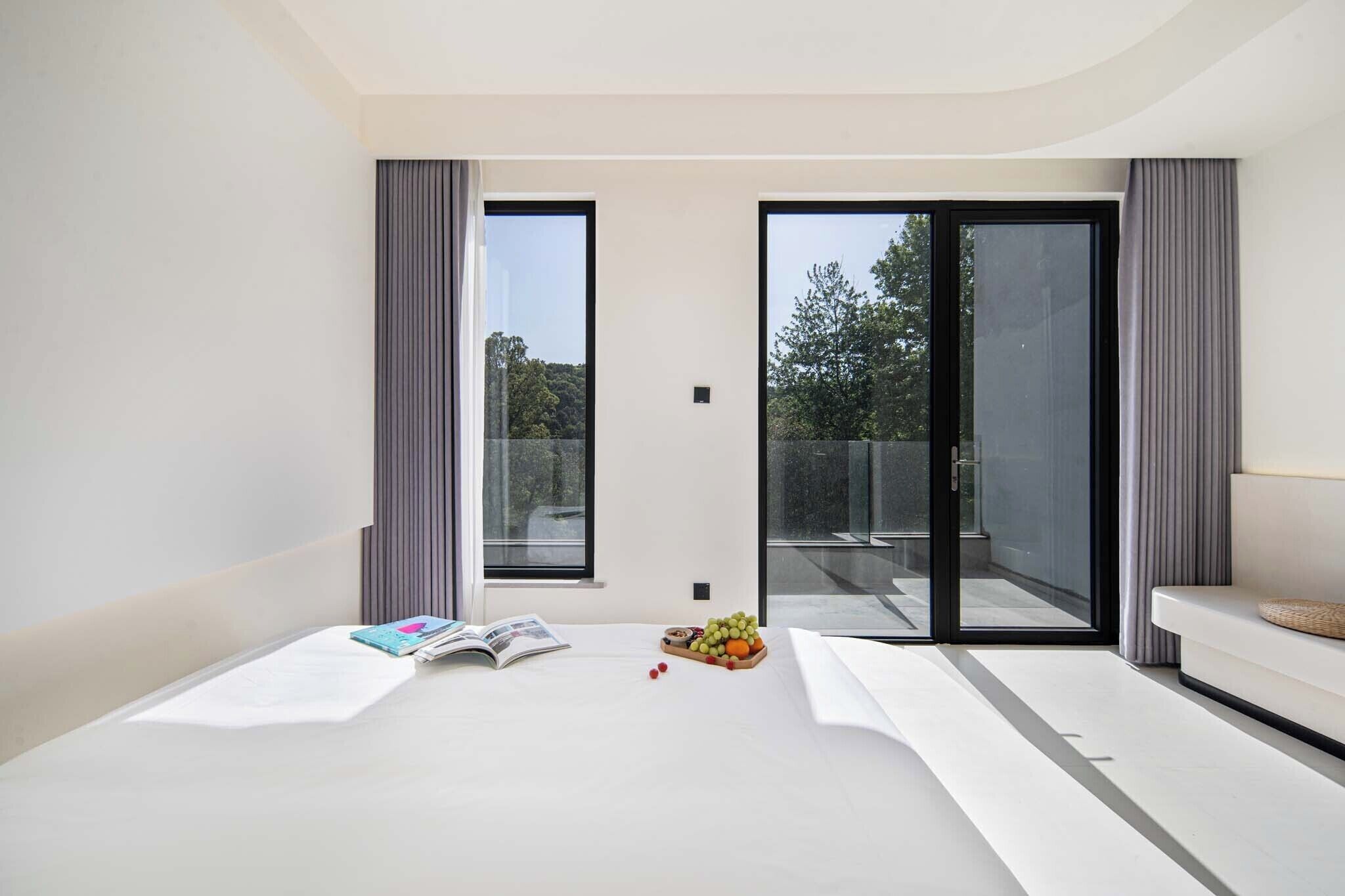 Quarto moderno com uma grande porta de correr de vidro, uma cama branca, uma mesinha com um livro e fruta, e um banco com almofada.