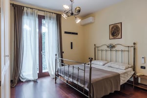 Quarto com estrutura de cama de metal, mesa de cabeceira, ar-condicionado fixado na parede, quadro emoldurado e janela com cortinas.
