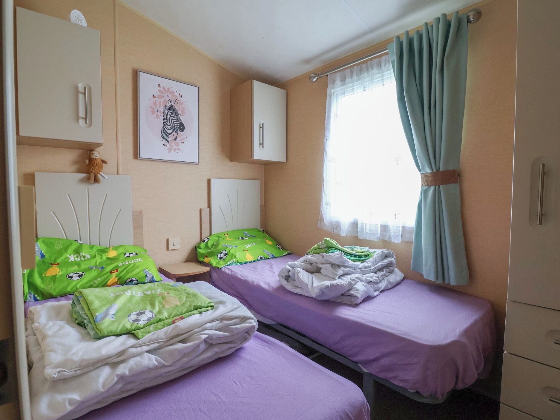 Quarto com duas camas, janela com cortinas, quadro pendurado na parede e um pequeno prateleira com um urso de pelúcia.