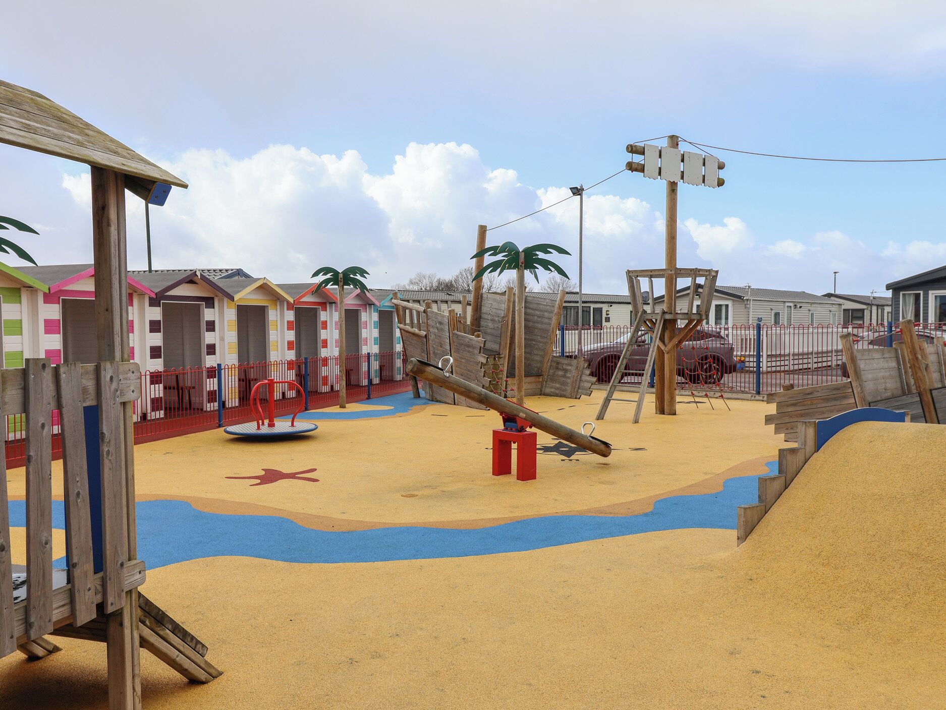 Uma área de recreação infantil com areia, gangorra e casinhas coloridas.