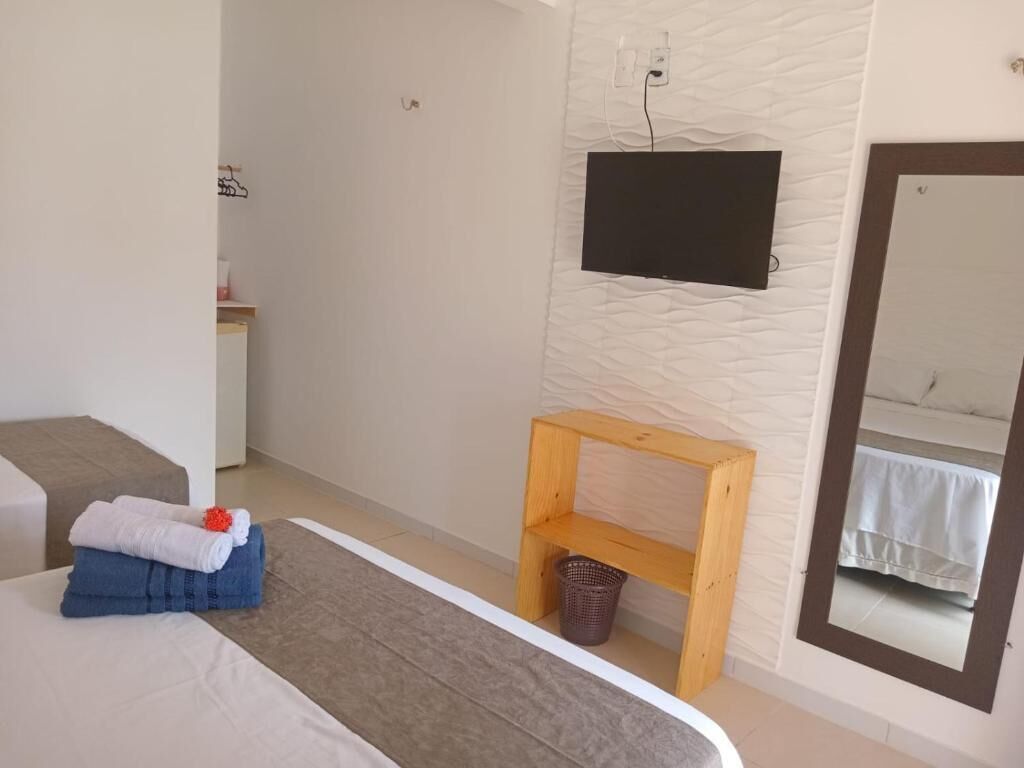 Quarto de hotel com cama, mesa de cabeceira de madeira, TV fixada na parede e espelho.