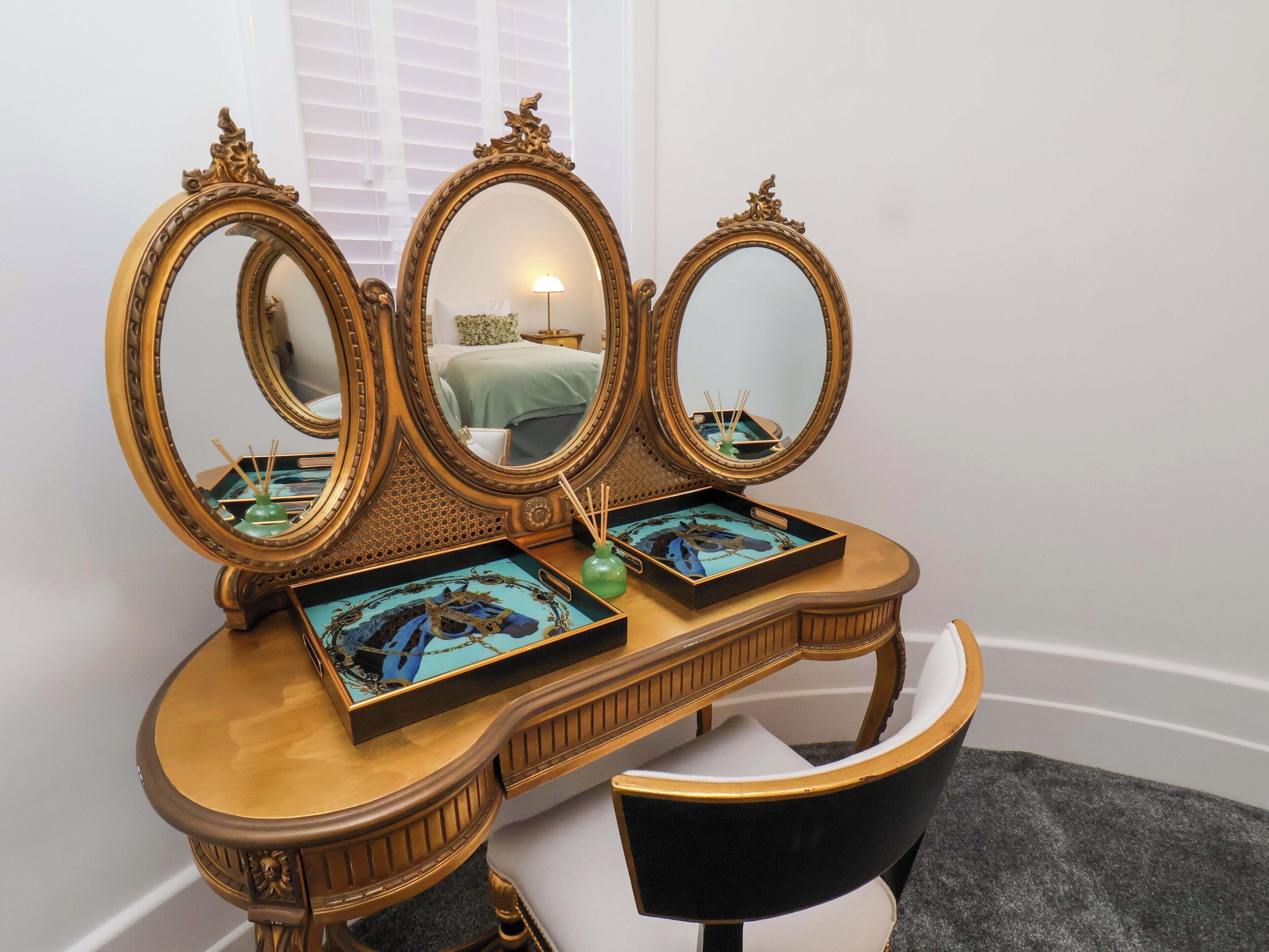 Uma toucador com três espelhos, um tabuleiro com objetos decorativos e uma cadeira.