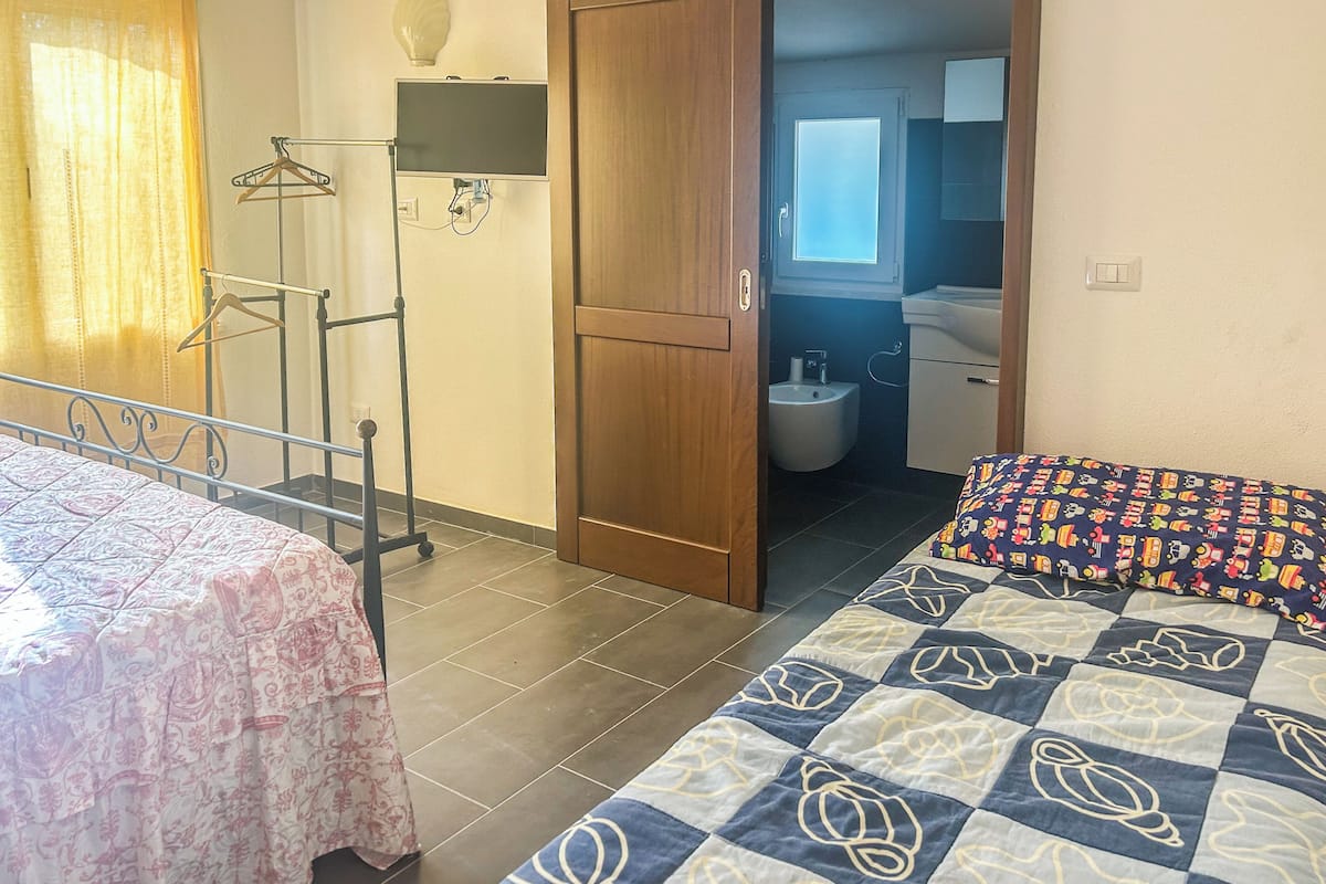 Quarto com cama, guarda-roupa, mesa de cabeceira, espelho e banheiro visível por uma porta aberta.