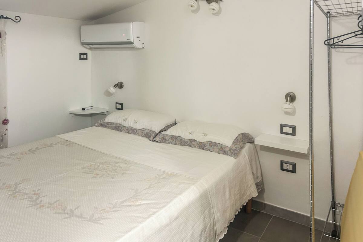 Quarto com cama, ar condicionado fixado na parede, mesa de cabeceira, luminária e varal de roupas.