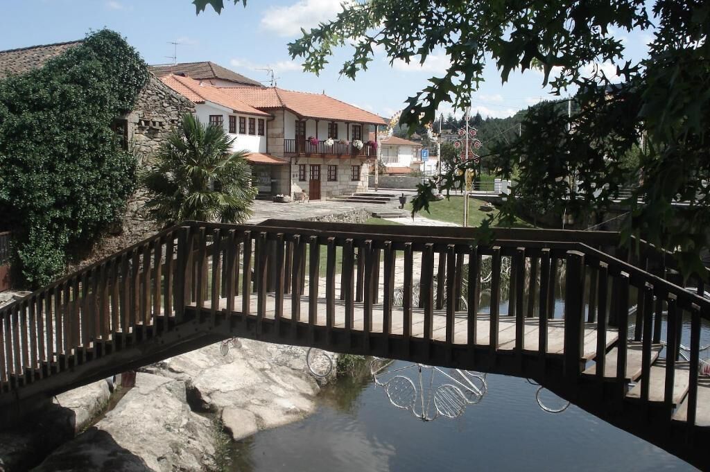 Uma ponte de pedra sobre águas calmas, um edifício de pedra com telhado de telhas vermelhas e vegetação exuberante.