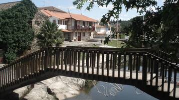 Uma ponte de pedra sobre águas calmas, um edifício de pedra com telhado de telhas vermelhas e vegetação exuberante.