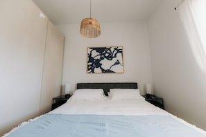 Quarto com uma cama, duas mesas de cabeceira, uma luminária pendente e um quadro abstrato na parede.