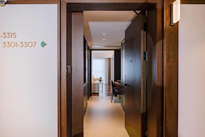 Corredor de hotel com portas de madeira, um balcão de recepção e vista para um quarto com cama e janela.