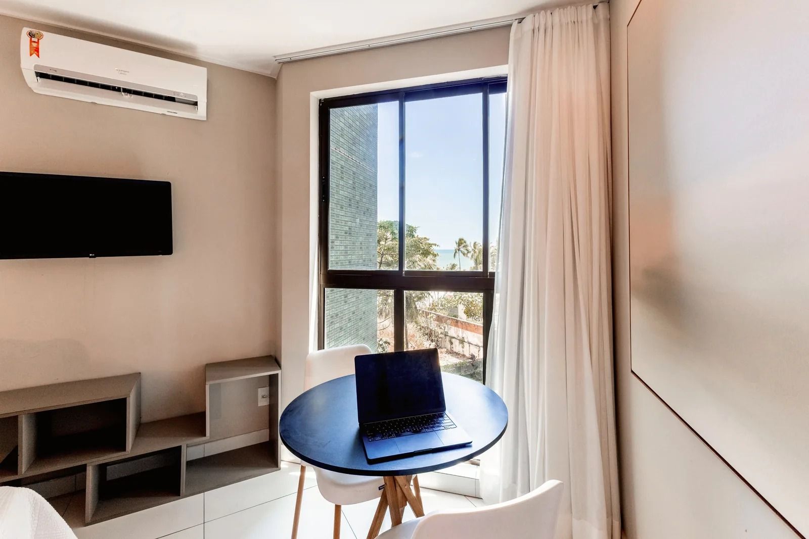 Quarto de hotel moderno com uma janela grande, TV de tela plana na parede, mesa redonda com um laptop e um pequeno espaço para sentar.