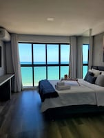 Quarto de hotel com uma janela ampla que oferece vista para o mar, uma cama com travesseiros e um cobertor, uma mesa de cabeceira com bandeja e um aparelho de ar condicionado.
