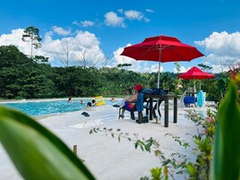 Uma área de piscina com superfície arenosa, guarda-sóis vermelhos e pessoas nadando e relaxando.