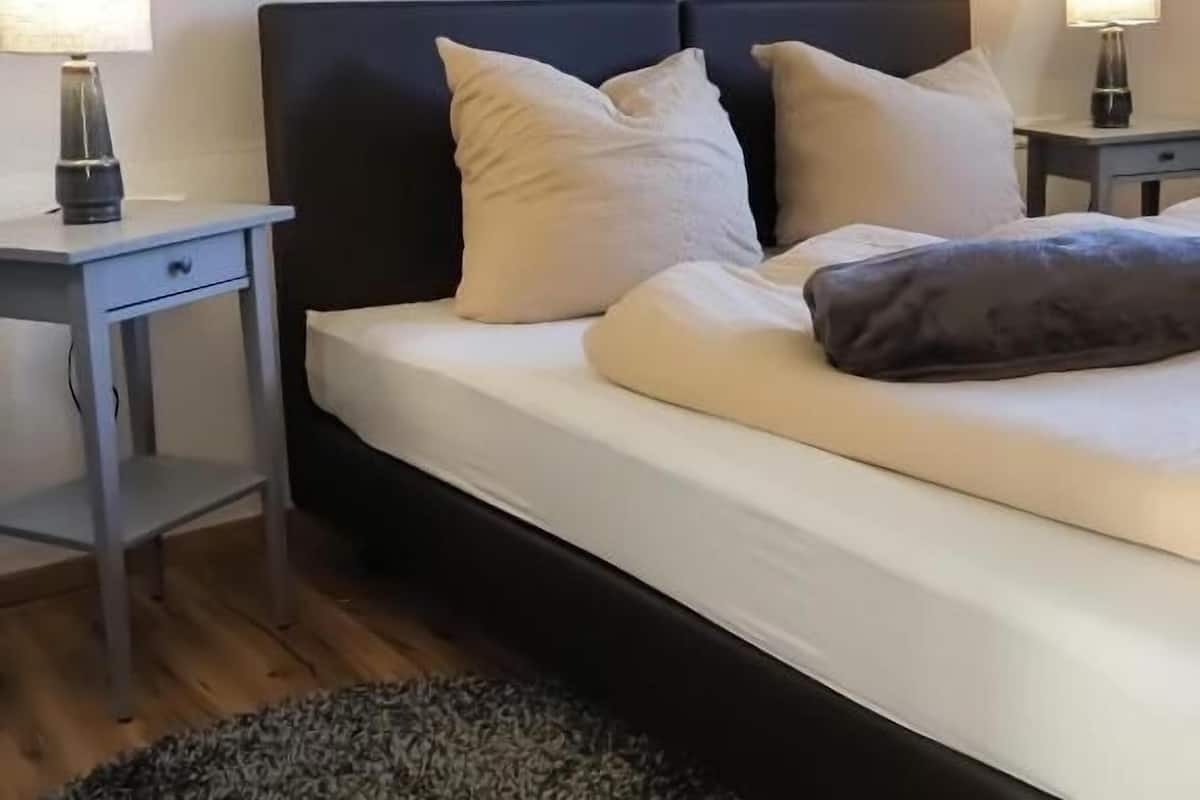 Cama bem arrumada com travesseiros e coberta, mesa de cabeceira com abajur e um tapete pequeno no chão.