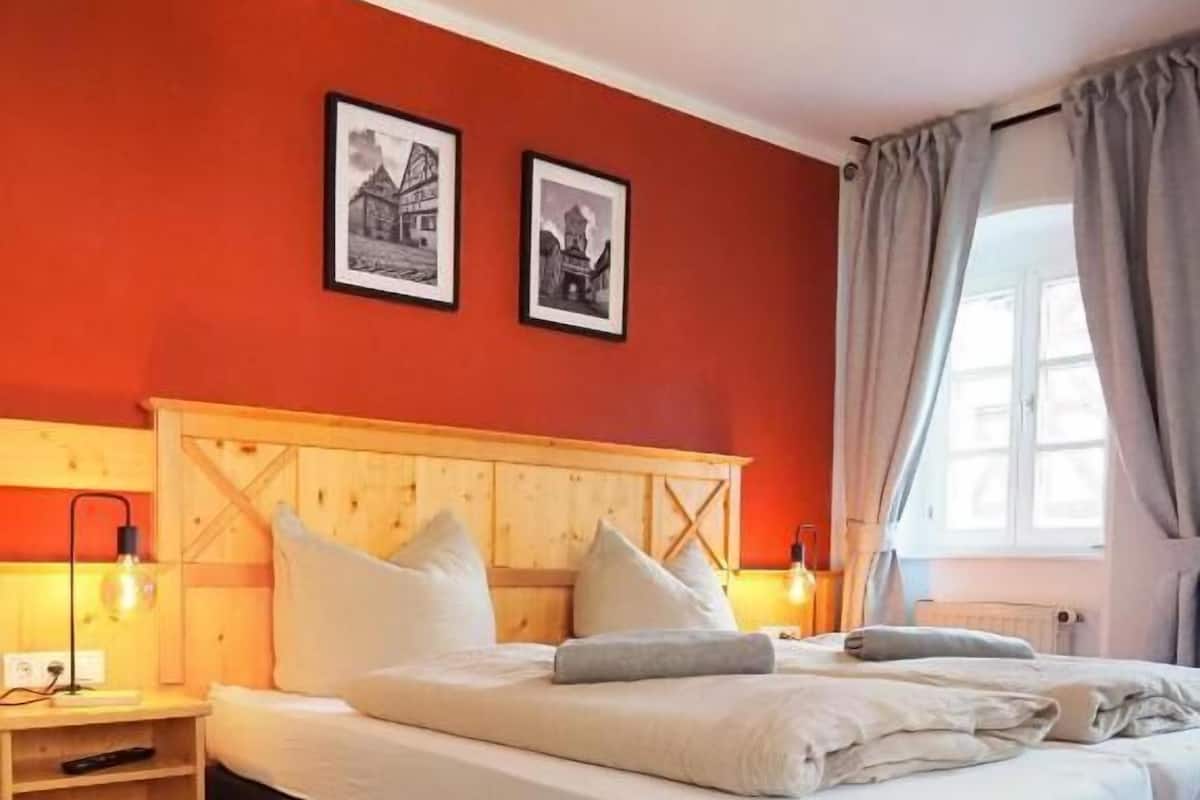 Quarto com cabeceira de madeira, cama com lençóis brancos, duas imagens emolduradas na parede e uma janela com cortinas.