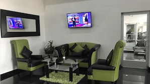Um lobby moderno com um sofá verde e preto, uma mesa de centro de vidro e duas televisões de tela plana fixadas na parede.