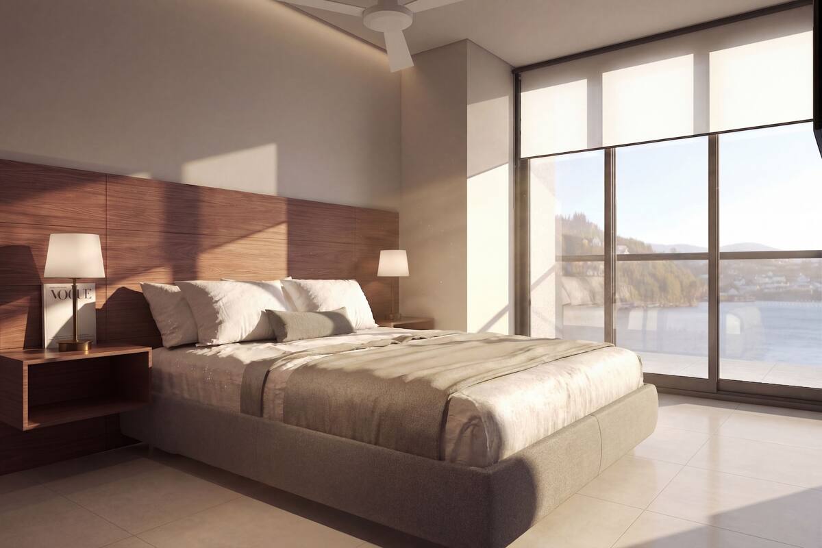 Quarto moderno com uma cama grande, mesas de cabeceira e vista para a água através de janelas amplas.