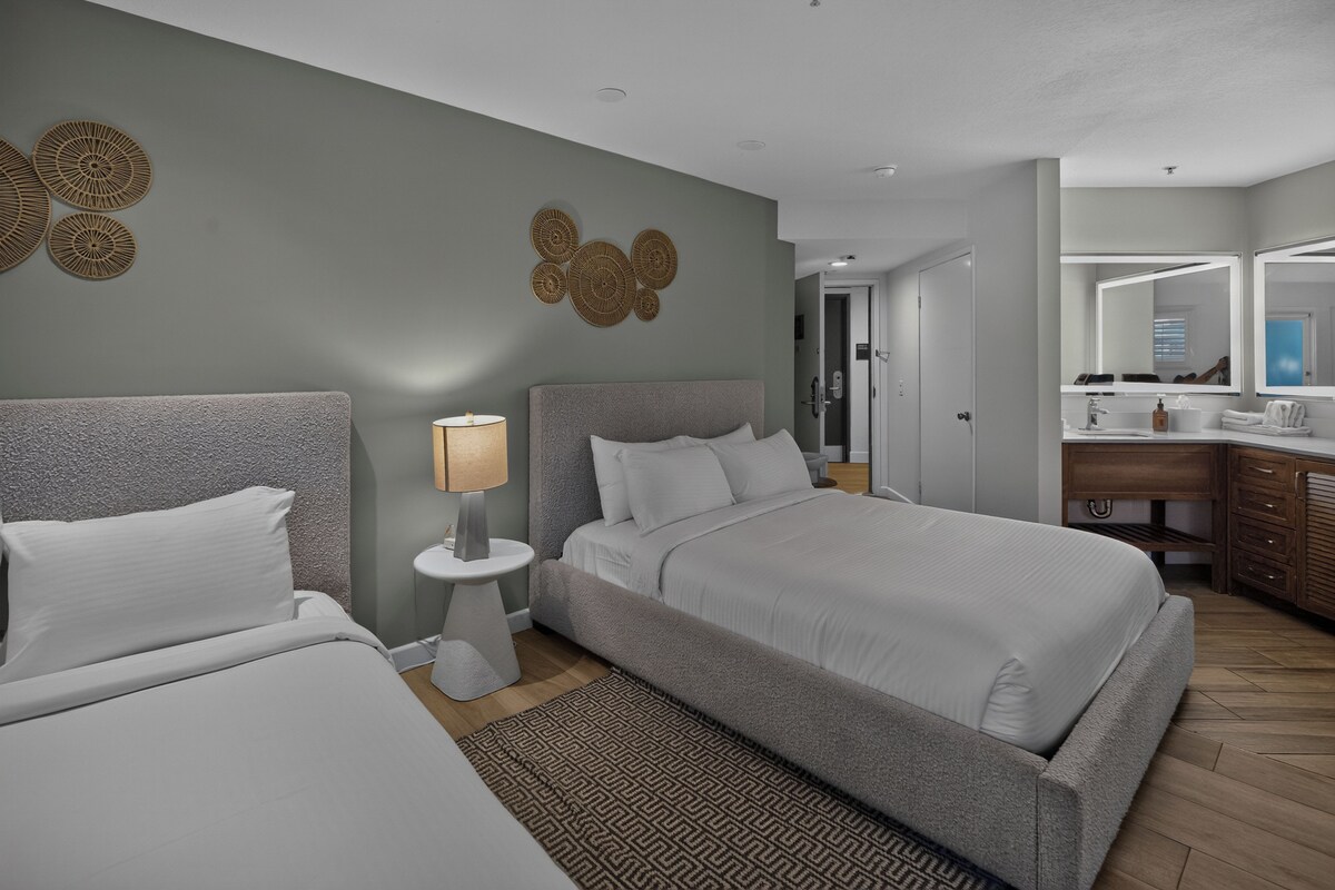 Quarto de hotel com duas camas, mesa de cabeceira com abajur, banheiro com espelho e tapete estampado.