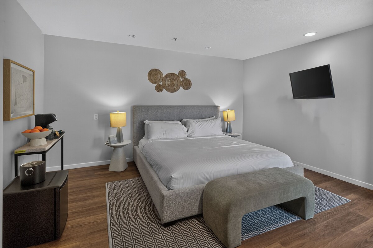 Quarto moderno com cama grande, luminárias de cabeceira, TV de tela plana e um detalhe decorativo na parede.