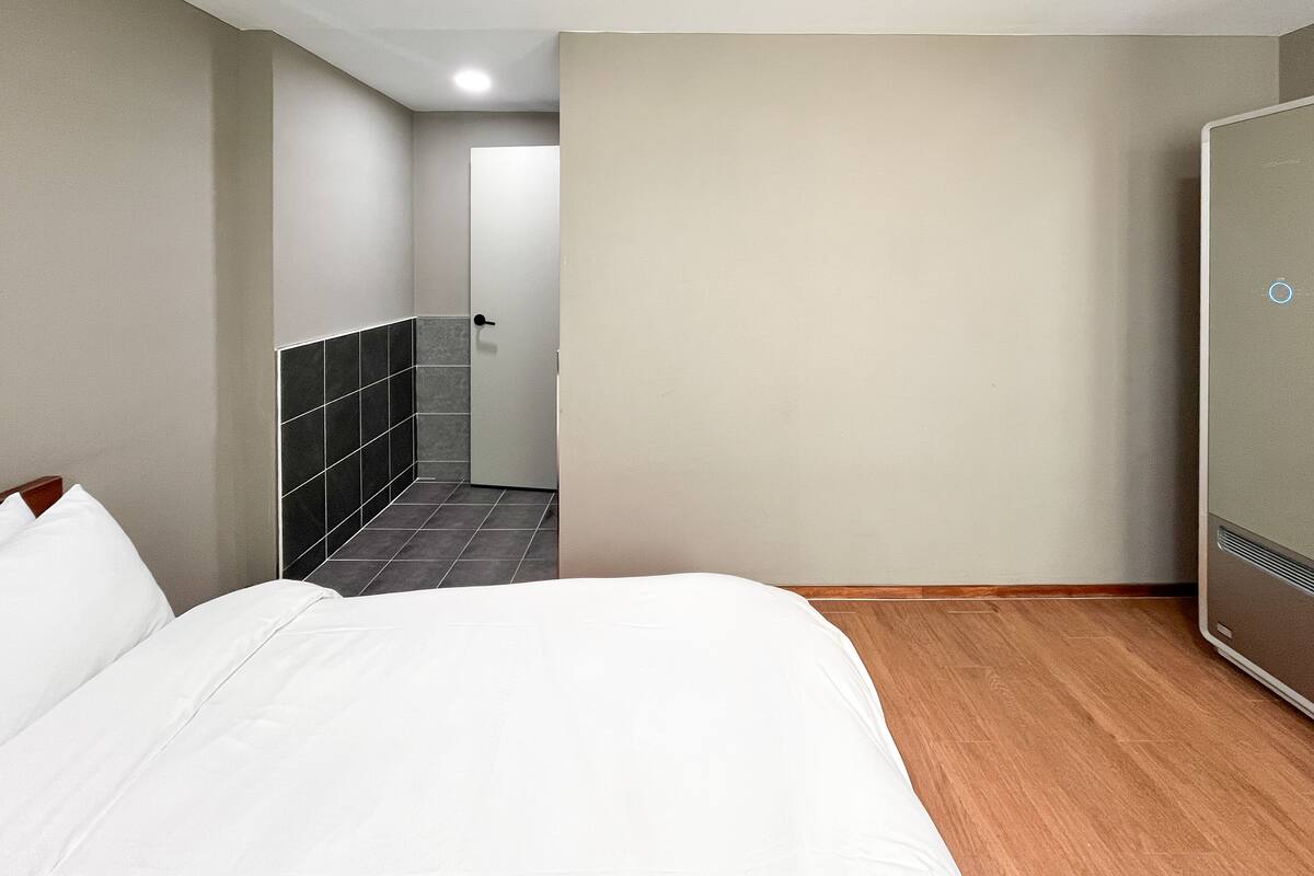 Quarto de hotel com cama, piso de madeira, área de chuveiro com azulejos escuros e porta branca.