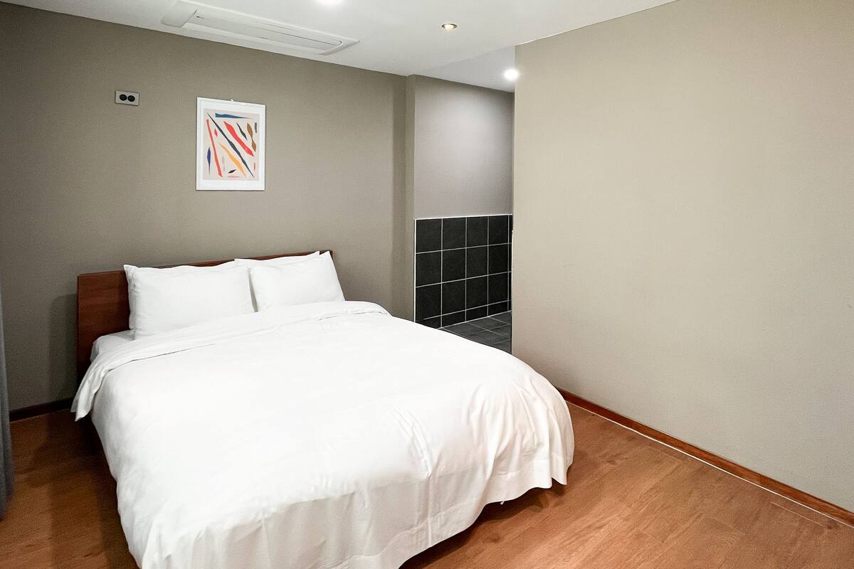 Cama bem arrumada com lençóis brancos, cabeceira de madeira e uma obra de arte abstrata na parede.