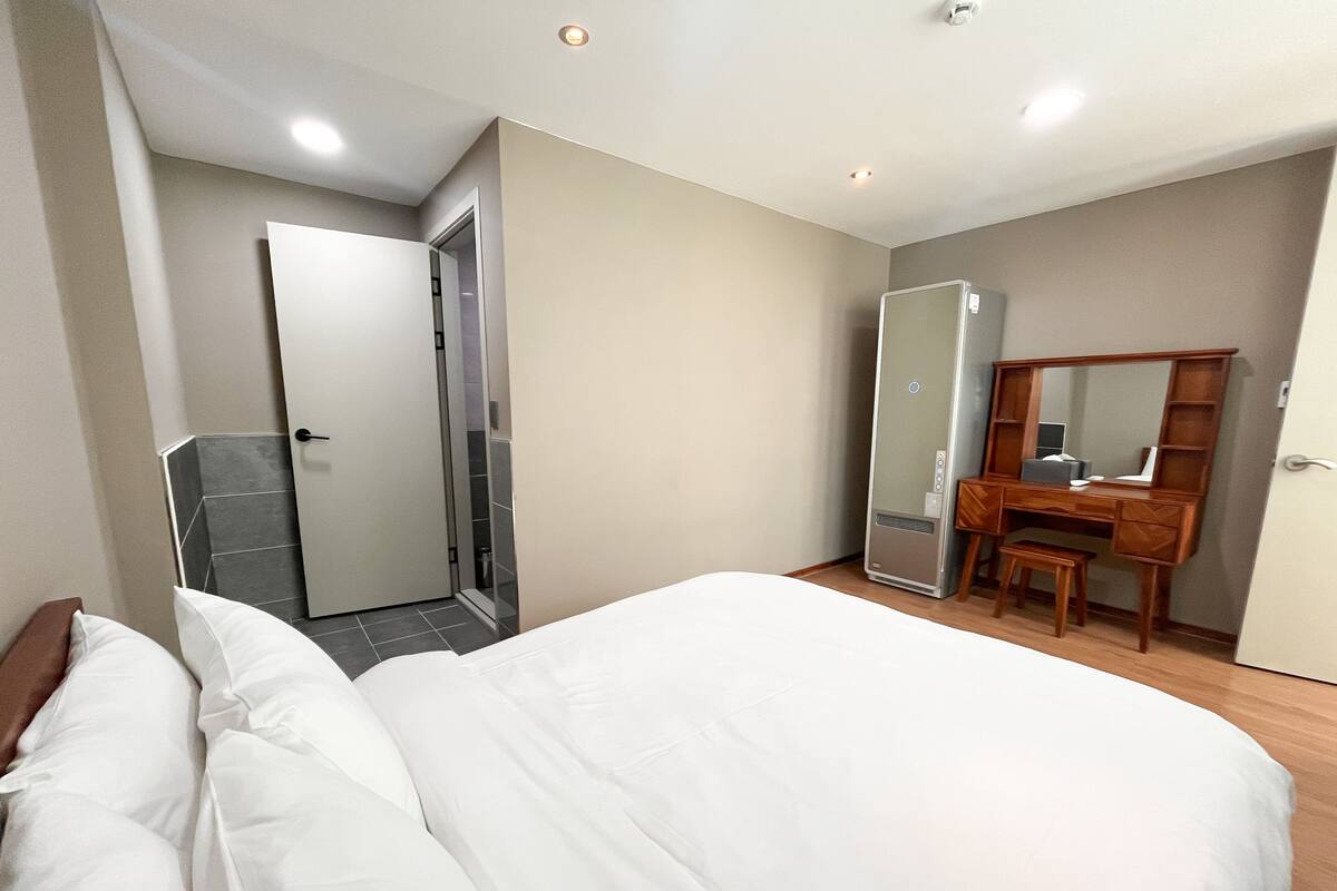 Quarto de hotel com cama, escrivaninha com espelho, guarda-roupa e chuveiro.