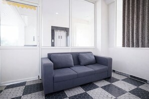 Quarto com sofá cinzento, uma janela grande com cortinas, chão em mosaico e porta branca com barra de empurrar.