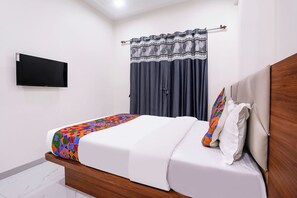 Uma cama bem arrumada com um cobertor colorido estampado com padrões geométricos, cabeceira de madeira, televisão de tela plana na parede e uma janela com cortinas escuras.