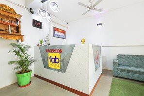 Uma mesa de recepção com uma placa 'fab hotels', um relógio, um ventilador de teto, uma planta verde e um sofá.