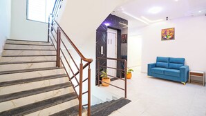 Uma escadaria com degraus de mármore e corrimãos de metal, que leva a um quarto com um sofá azul, uma planta em vaso e uma porta com um design de parede singular.
