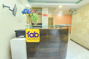 Uma receção com bancada de vidro, o letreiro 'fab hotels', um vaso de flores e uma área de receção com um pilar de pedra e uma porta ao fundo.