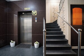 Um elevador com o indicador de pavimento 'G', uma escada com corrimão de metal e duas plantas em vasos no chão.