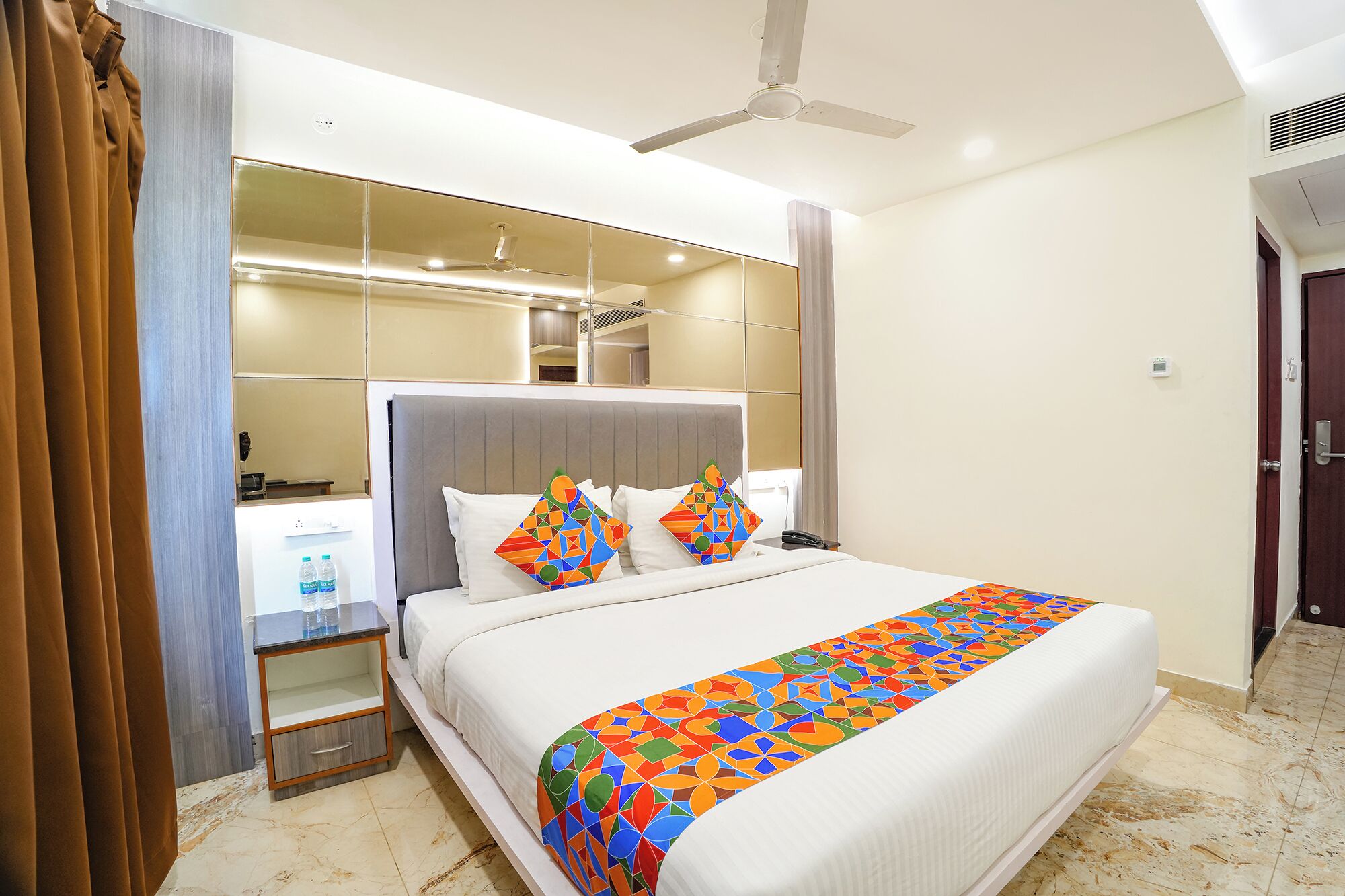 Quarto de hotel com cama, mesa de cabeceira, espelho, ventoinha no teto e porta.