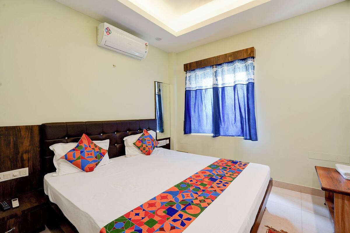 Quarto de hotel com cama, mesa de cabeceira, escrivaninha, janela com cortinas e ar condicionado.