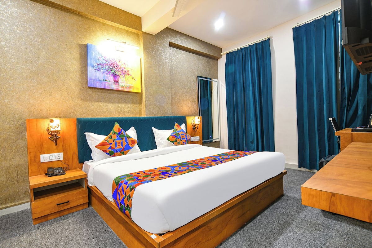 Quarto de hotel com uma cama grande, uma mesinha de cabeceira de madeira, uma escrivaninha, um quadro na parede e cortinas azuis.