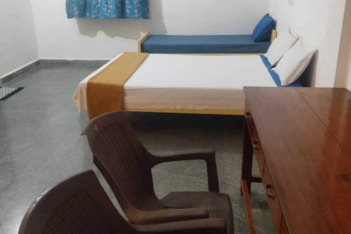 Quarto pequeno com uma cama de solteiro, uma mesa de madeira e duas cadeiras de plástico.