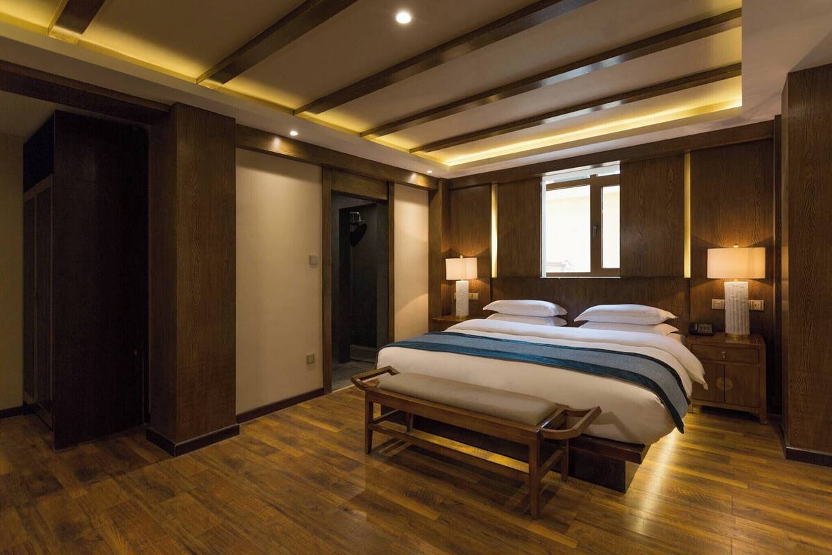 Quarto moderno com cama grande, luminárias de cabeceira e um banquinho de madeira.