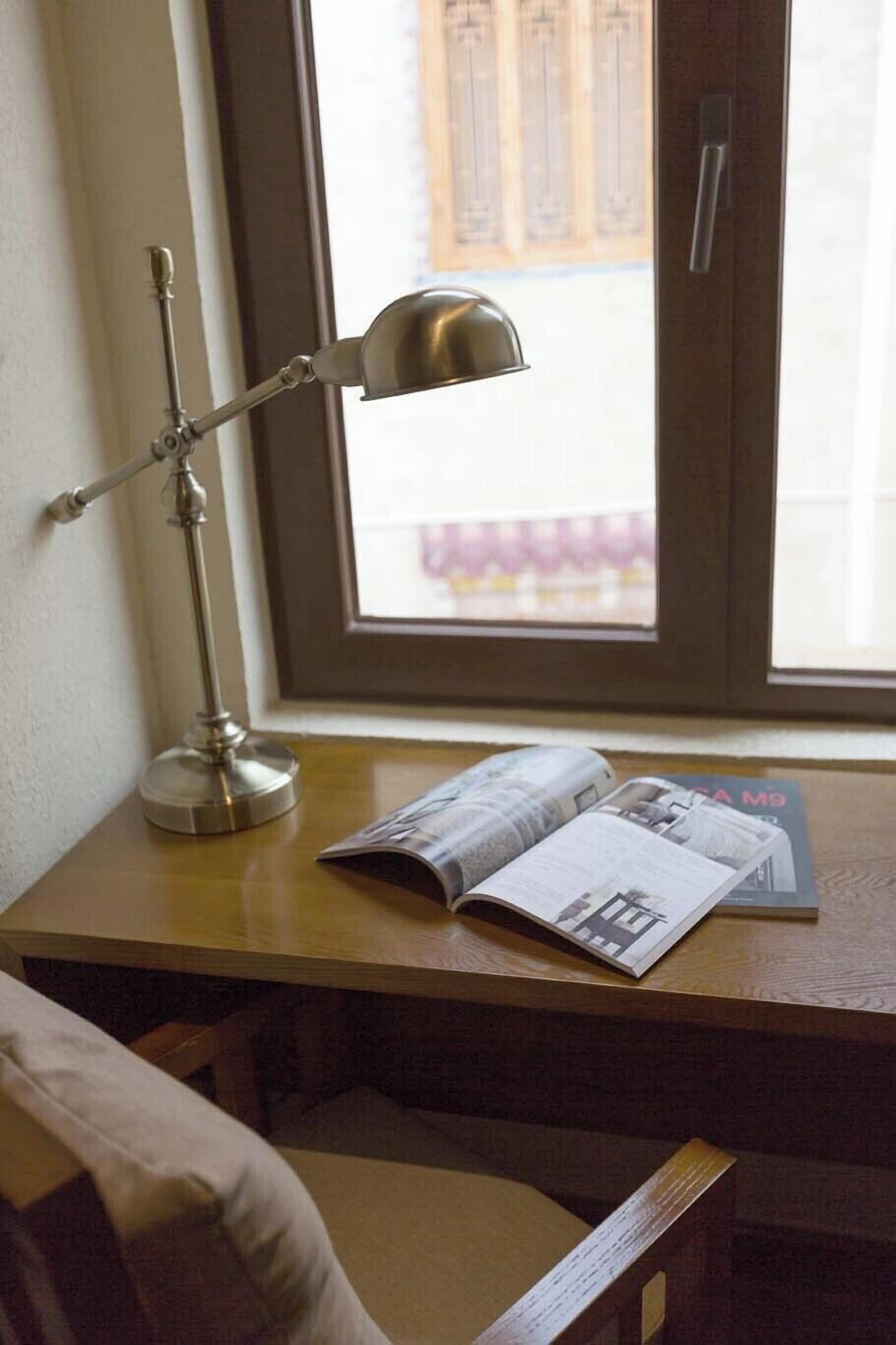 Uma mesa com uma luminária, uma revista e uma janela com vista para edifícios.