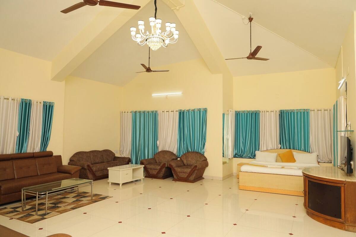 Uma sala de estar ampla com teto alto, lustre, ventiladores de teto, sofá, poltronas, mesa de centro e cortinas.