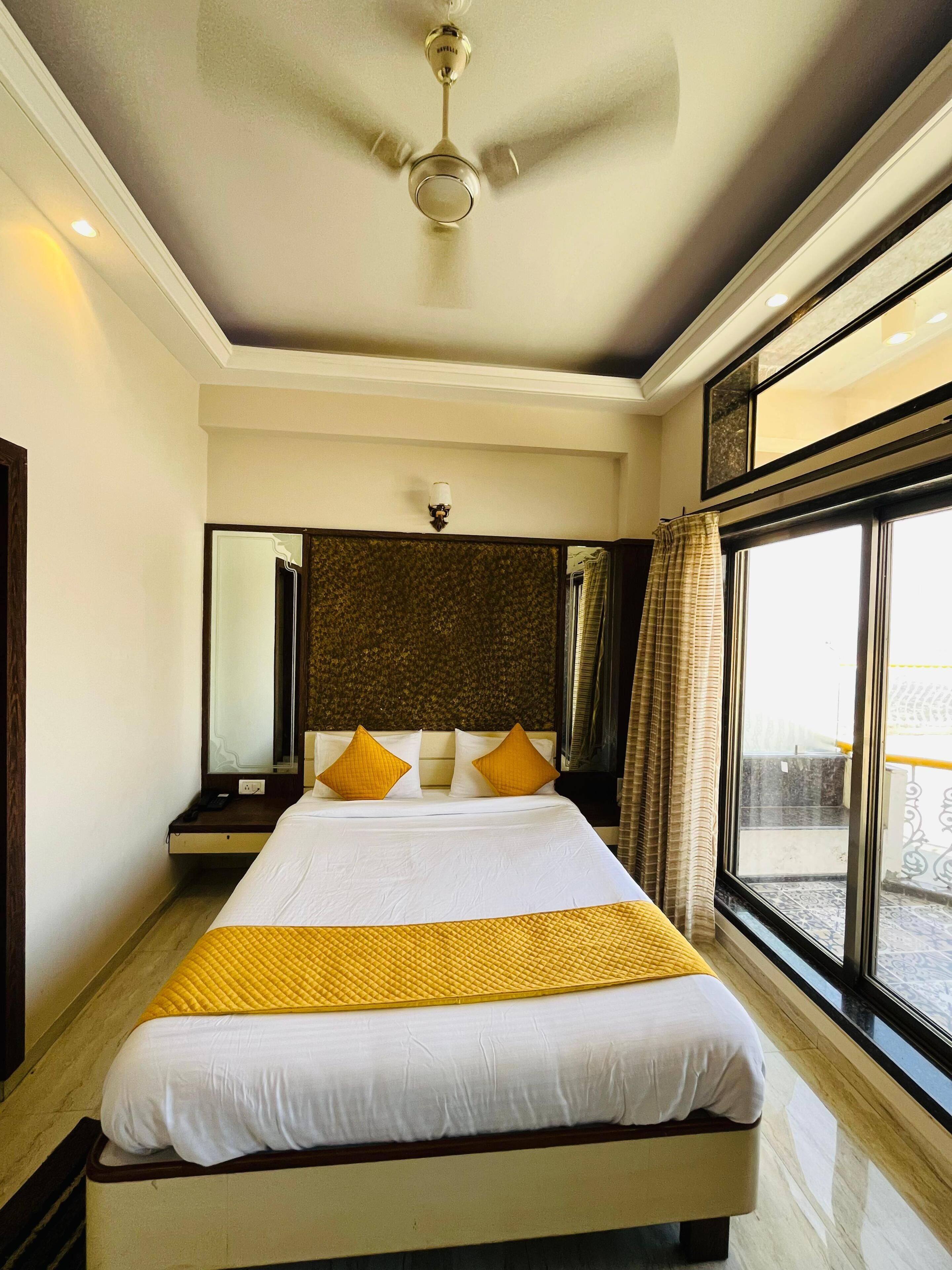 Quarto de hotel com cama, ventoinha no teto, uma janela grande e um espelho.
